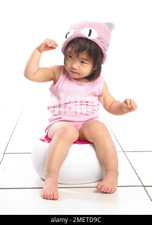 Girl wearing pink vest and pink wool hat, red slippers sitting on white potty holding nappy, 20 ...