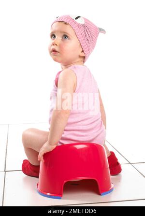 Girl wearing pink vest and pink wool hat, red slippers sitting on white potty holding nappy, 20 ...