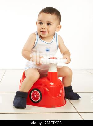 Boy wearing vest and slippers sitting on red car potty with steering wheel, 15 months Stock ...
