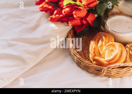 Wicker tray with pastries and coffee and flowers on white bed linen ...