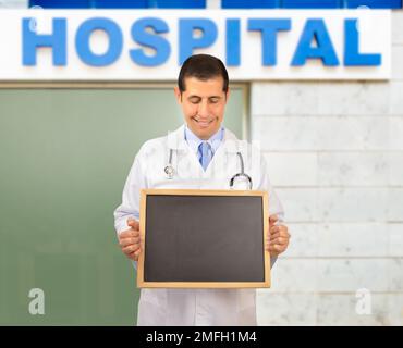 Doctor standing and smiling with chalkboard at hospital with copy space ...