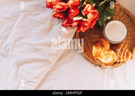 Wicker tray with pastries and coffee and flowers on white bed linen ...