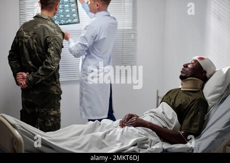 Military man with bleeding wound in head lying on bed in hospital ward ...