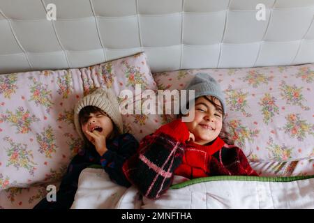 Girls wearing warm hats lay in the bed under blanket hugging each other ...