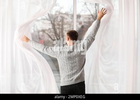 Young man opening window curtains at home, back view Stock Photo - Alamy