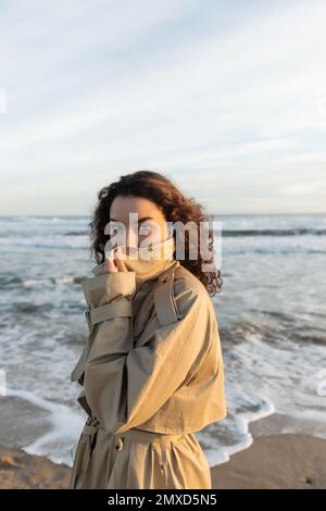 curly young woman adjusting wired earphones while holding smartphone ...