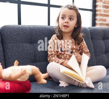 Adorable hispanic girl reading book sitting on sofa at home Stock Photo ...