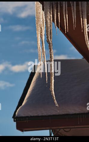 A closeup of ice dams hanging from a roof with cloudy sky blurred ...