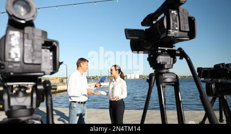 Journalist interviewing young woman near river, focus on video cameras ...