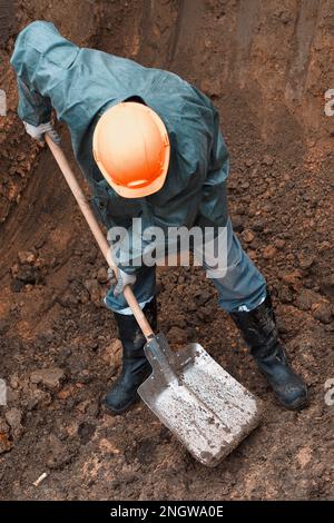 Man in helmet, raincoat and work clothes with shovel in pit digs hole ...