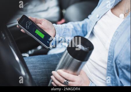 Woman with thermos flask using electric car charging app seen through ...