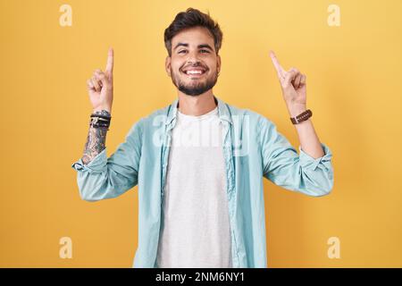 Hispanic man with tattoos standing over blue background pointing aside ...