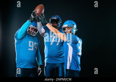 Three American football players raising their hands up holding the ball ...