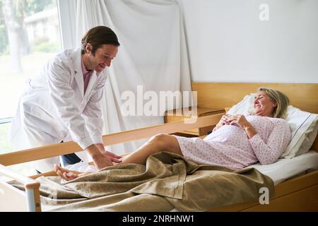 Smiling male doctor lifting legs of senior man lying in bed at ...