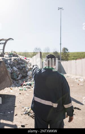 Landfill worker directing skid steer loader on the garbage heap, rear ...