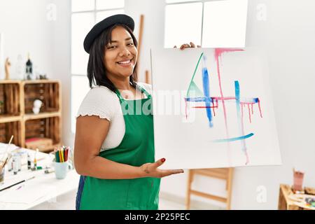 Hispanic brunette woman holding canvas proud of painting at art studio ...