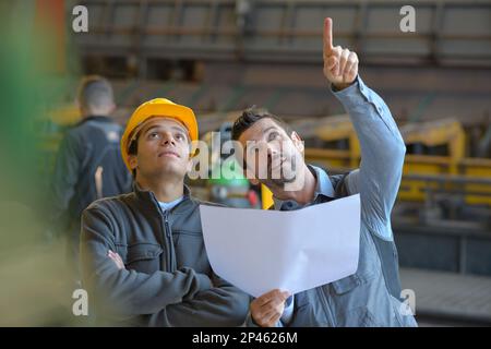Two factory workers pointing upwards while discussing together in ...