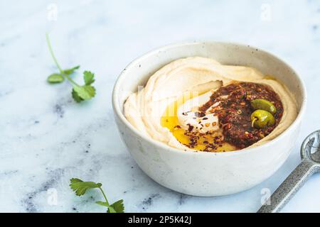 Homemade spicy hummus with tahini and olive oil in a bowl, white marble ...