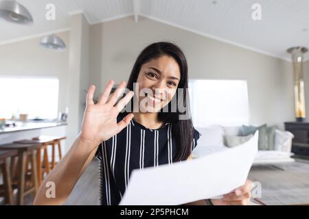 Woman having video call and waving, enjoying coffee in modern home ...