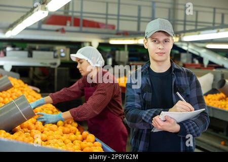 Young foreman with papers controlling work on tangerines sorting line ...