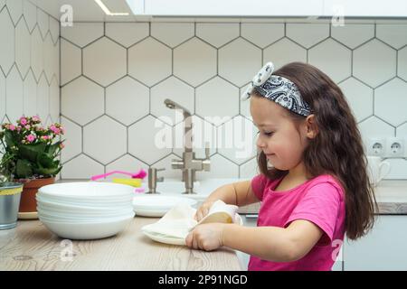 Happy little girl wiping clear plates with dry towel on kitchen set ...