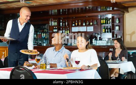 Polite waiter serving pizza to hispanic couple in restaurant Stock ...