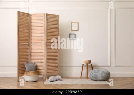Folding screen, small table and knitted pouf near white wall in room ...