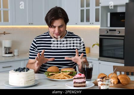 Hungry overweight man at table with sweets and fast food in kitchen ...