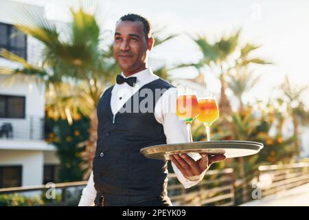 Holds cocktails. Black waiter in formal clothes is at his work outdoors ...