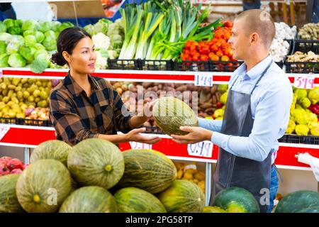 Greengrocer worker explaining woman how to choose melon Stock Photo - Alamy