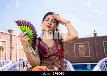 Exhausted Indian woman using a paper hand waver fan sitting outdoor in ...