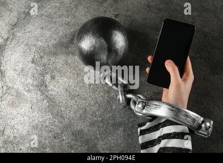 Man shackled with ball and chain holding smartphone at grey table, top ...