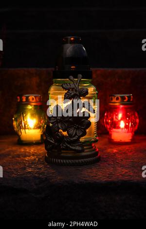 Different grave lanterns with burning candles on granite tombstone outdoors. Funeral ceremony ...