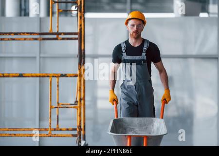 Walks with wheelbarrows. Man in grey uniform works indoors in modern big office at daytime Stock ...