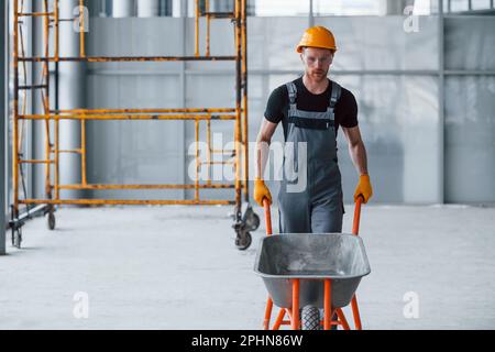Walks with wheelbarrows. Man in grey uniform works indoors in modern ...