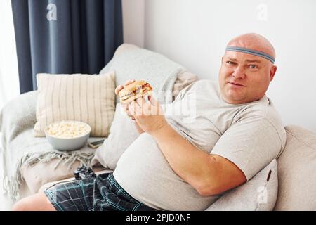 Sits on the sofa with popcorn and hamburger. Funny overweight man in ...