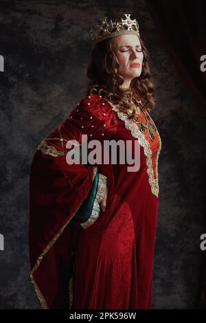 tired medieval queen in red dress with crown having backache on dark ...