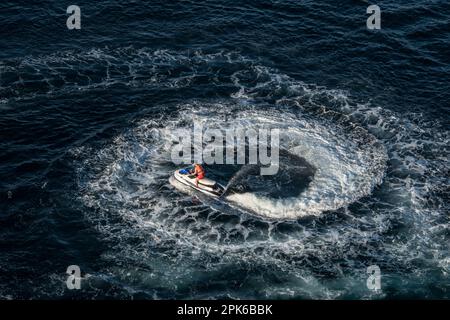 An unidentifiable man rides a personal watercraft, jet ski, creating a ...