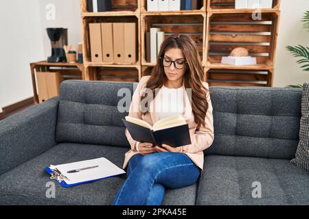 Young hispanic woman psychologist reading book and writing on clipboard ...