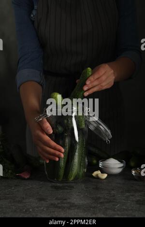 Woman putting cucumber into glass jar at white marble kitchen table ...