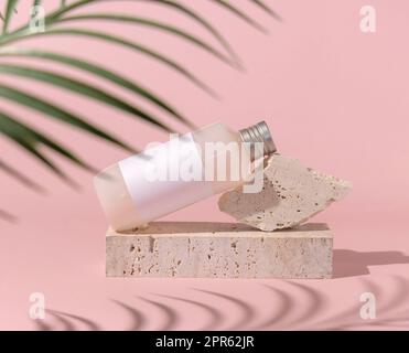 Cosmetic bottle with blank label lies on stone on light pink, palm leaf ...