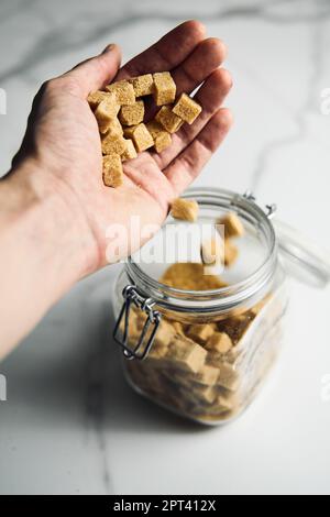Brown sugar cubes in a man's hand and in the jar on white marble ...