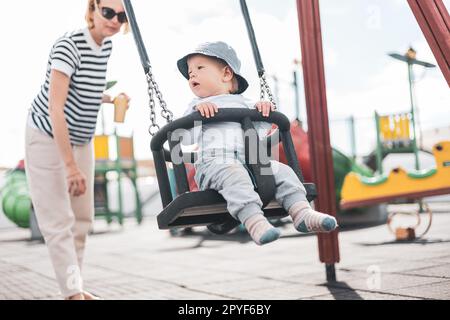 Mother pushing her infant baby boy child on a swing on playground ...