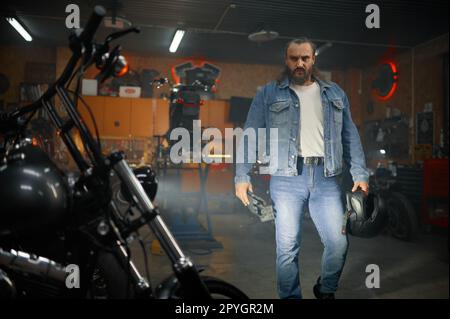 Portrait of bearded brutal biker looking motorcycle in garage workshop ...