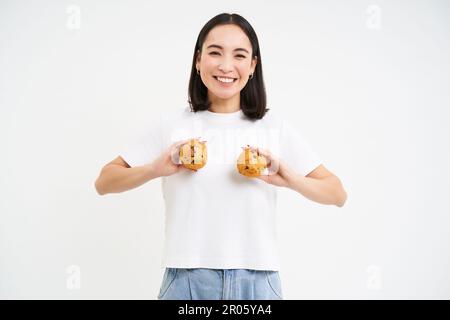 Happy japanese woman shows cupcakes on chest, eating pastry, standing ...