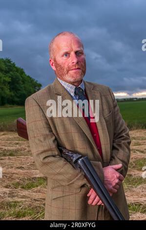 An English gentleman stood at twilight in a tweed shooting suit with a ...