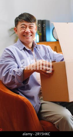 Smiling Asian guy unpacking online shopping at home, holding hoodie ...