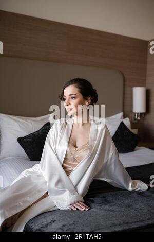 brunette bride in silk robe smiling at camera while touching face in ...
