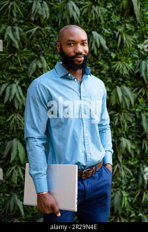 Bearded businessman holding laptop standing near panoramic window in ...
