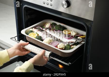 Woman putting baking tray with fish and vegetables into oven Stock ...